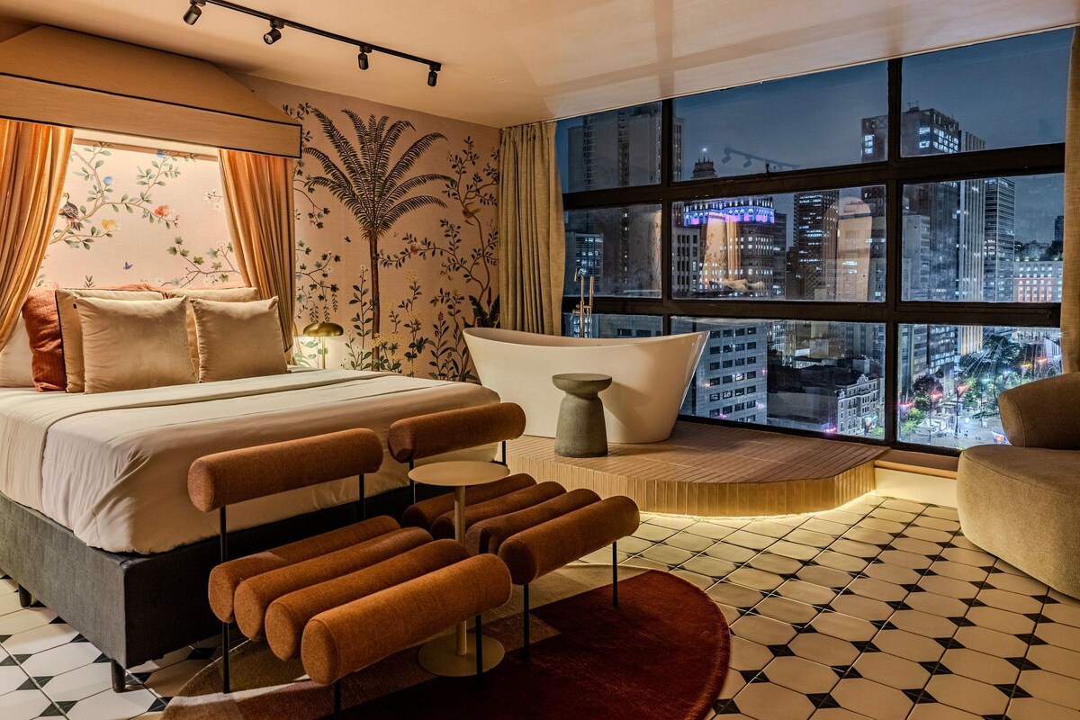 Quarto de hotel moderno com uma cama grande, um espaço de estar com bancos, uma mesa pequena e vista para a cidade através de grandes janelas.