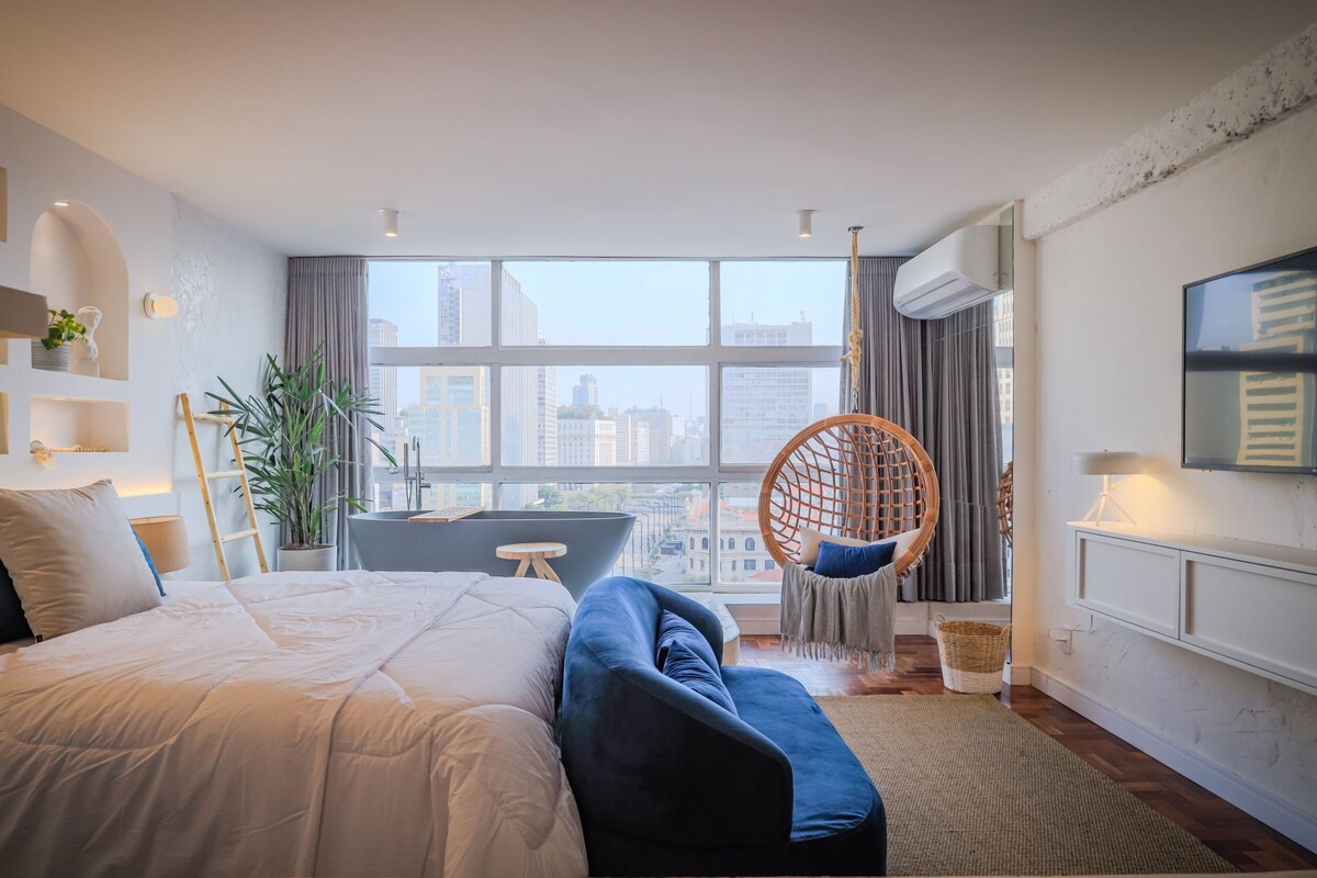 Quarto de hotel moderno com uma cama grande, um sofá azul, uma cadeira suspensa, uma escrivaninha e vista para a cidade.