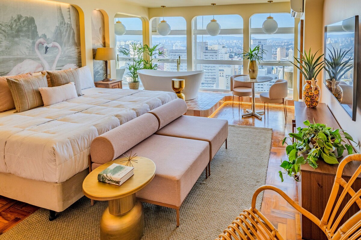 Quarto de hotel moderno com uma cama grande, área de estar, banheira e mesa de jantar com cadeiras.