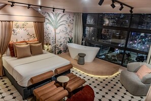 Quarto de hotel moderno com uma cama grande, área de estar com sofás, banheira e vista para a cidade através de janelas amplas.