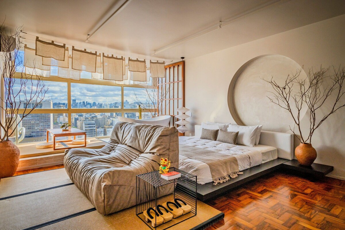 Uma sala moderna com sofá, cama, mesa de centro de madeira e uma janela grande com vista para a cidade.