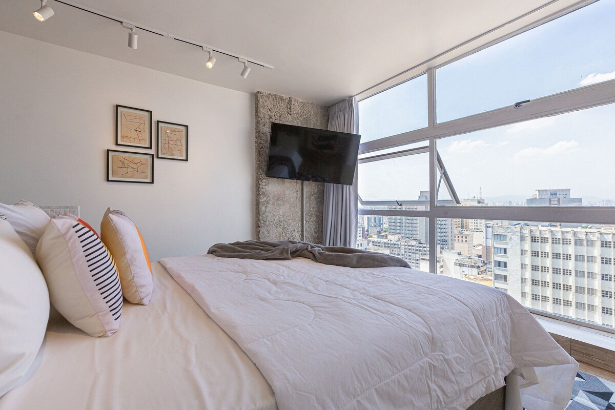 Um quarto moderno com uma cama grande, uma TV de tela plana presa a uma parede texturizada e vista para a cidade através de grandes janelas.
