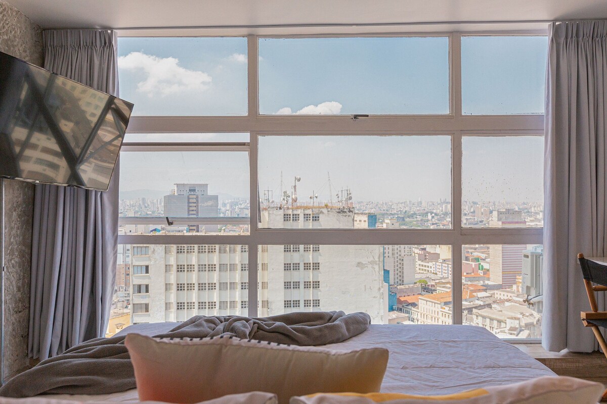 Quarto de hotel com uma janela grande, uma cama, uma TV de tela plana e uma cadeira.