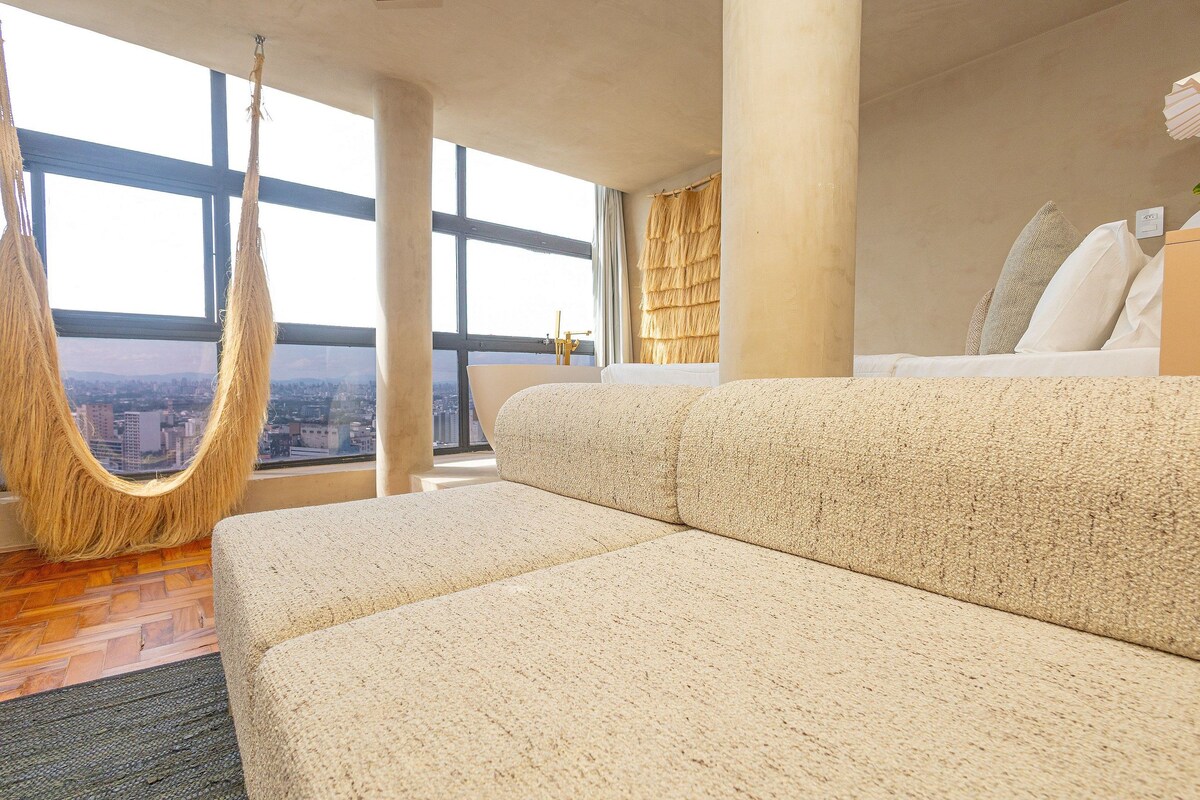 Quarto moderno com uma janela grande, uma cadeira suspensa, um sofá e uma cama com almofadas.