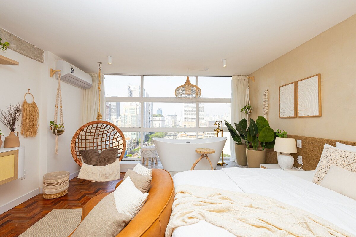 Quarto moderno com uma cama grande, um espaço de estar com uma poltrona, uma banheira e vista para a cidade.