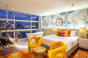 Quarto de hotel moderno com uma cama grande, duas cadeiras amarelas, uma cadeira suspensa, vista para a cidade e um mural tropical.