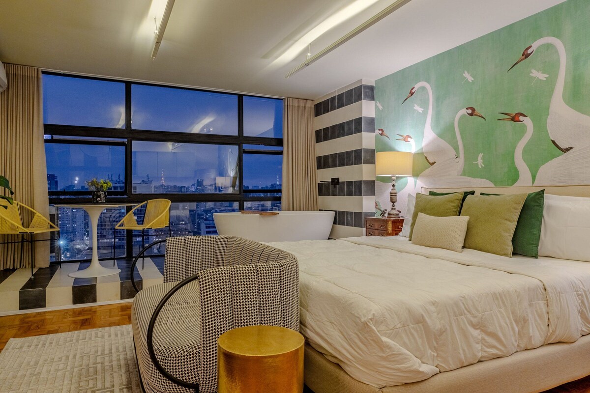 Quarto de hotel moderno com uma cama grande, vista para a cidade, uma mesa de jantar e uma cadeira.