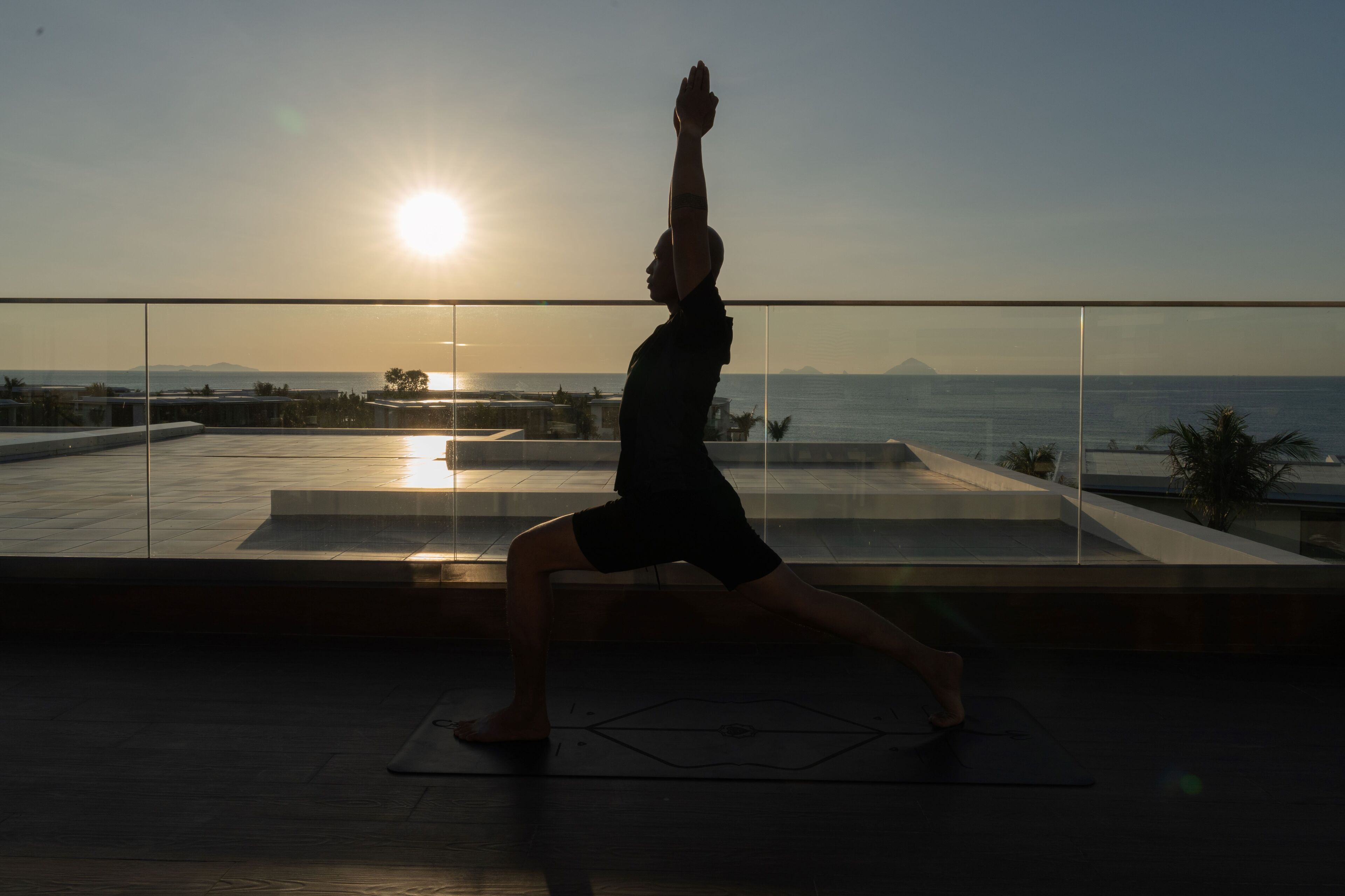 Uma pessoa fazendo yoga num tapete, com vista para o oceano e um pôr do sol ao fundo.