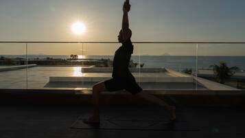 Uma pessoa fazendo yoga num tapete, com vista para o oceano e um pôr do sol ao fundo.