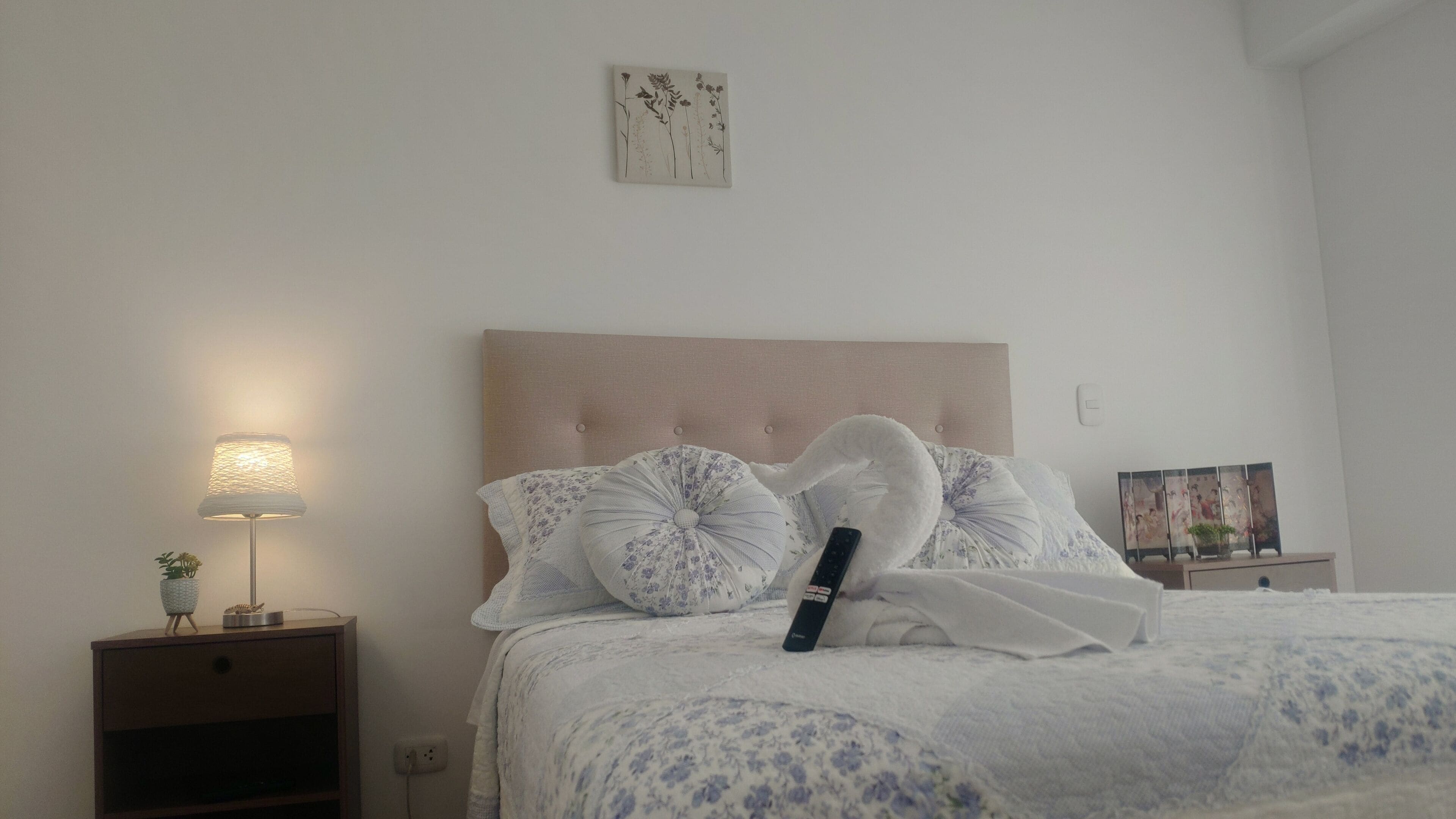 Uma cama bem arrumada com roupa de cama floral, cabeceira, abajur e uma pequena mesa com uma planta.