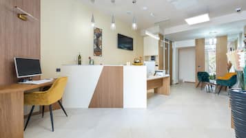 Uma área de recepção moderna com uma mesa de madeira, um computador e uma cadeira amarela. Há duas luminárias pendentes acima da mesa e uma janela grande ao fundo.
