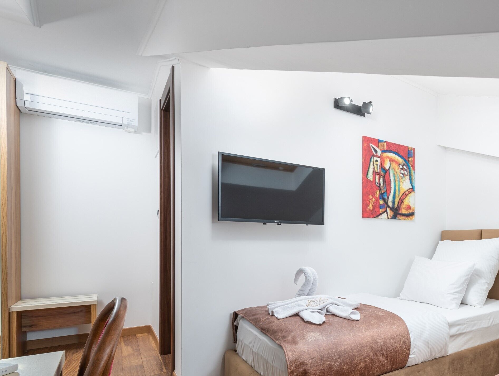 Quarto de hotel com cama, televisão na parede, um quadro e uma cadeira de madeira.