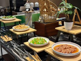 Disposição de buffet com diversos pratos, uma cesta de vime, uma planta em vaso e uma pequena mesa com um pote branco.