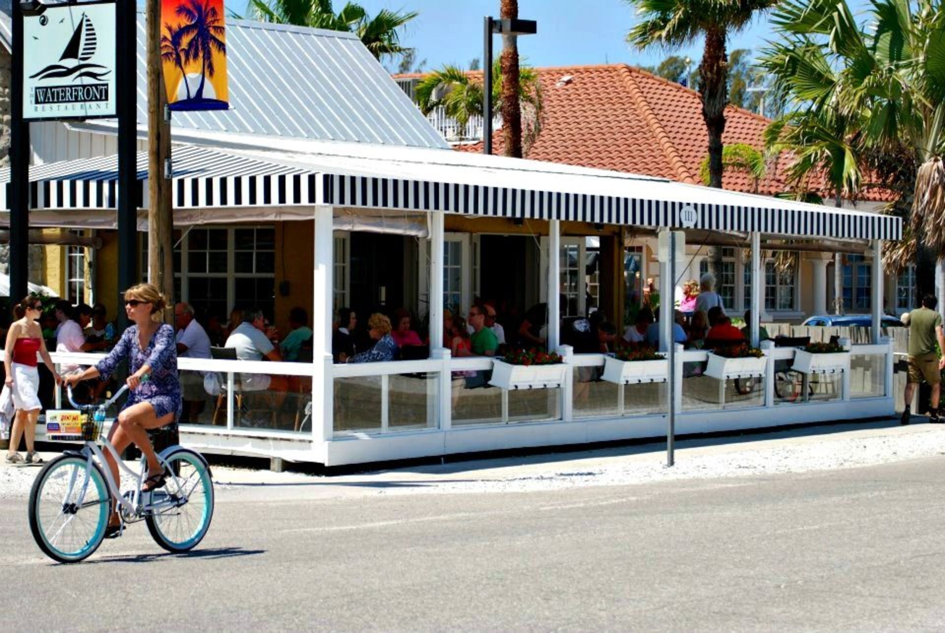 Restaurante à beira-mar com mesas ao ar livre, um ciclista passando e palmeiras ao fundo.