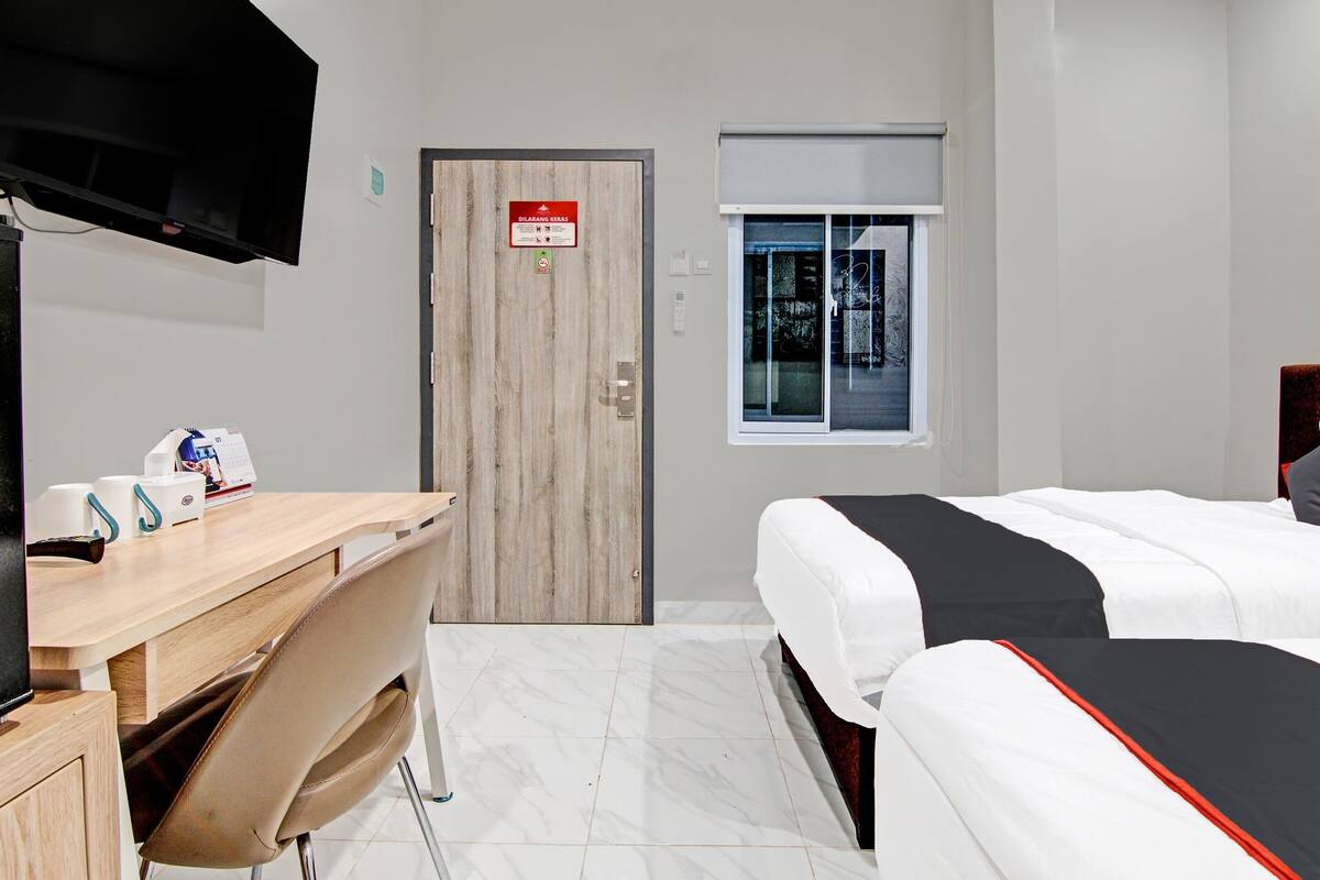 Quarto de hotel com porta de madeira, cama com roupa de cama preta e branca, escrivaninha com cadeira, televisão e janela com vista para árvores.