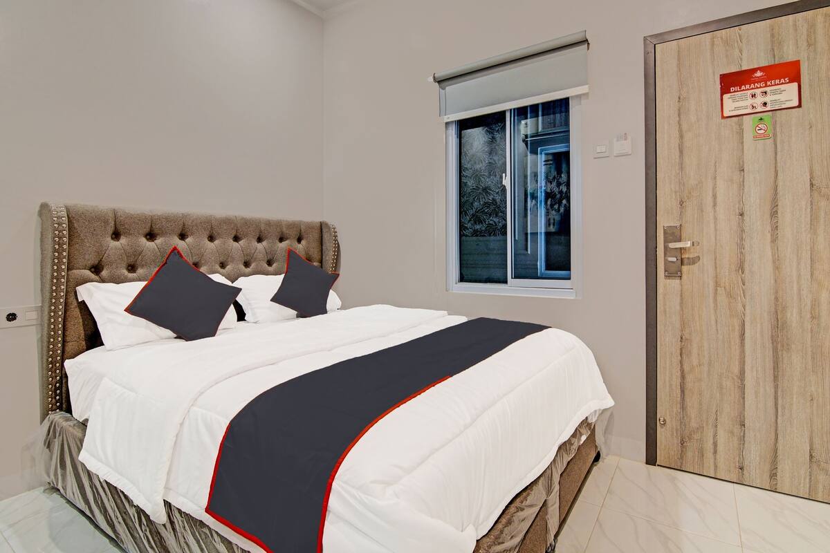 Cama bem arrumada com cabeceira estofada, janela com vista e porta com uma placa.