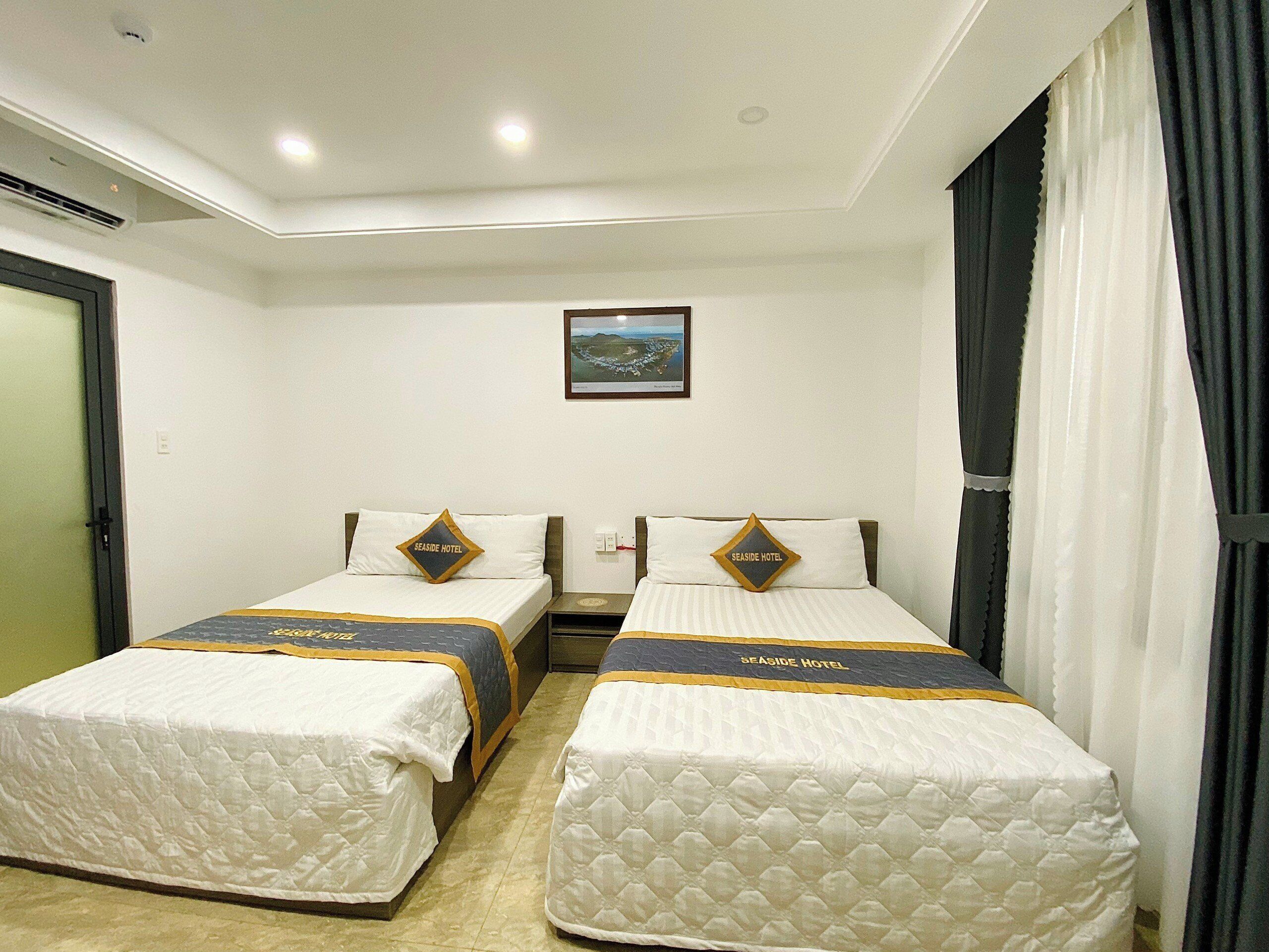 Quarto de hotel com duas camas, cada uma com colchão acolchoado e almofada com o logotipo do hotel. Há uma moldura com uma imagem na parede acima das camas, e uma janela com cortinas à direita.