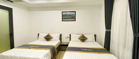 Quarto de hotel com duas camas, cada uma com colchão acolchoado e almofada com o logotipo do hotel. Há uma moldura com uma imagem na parede acima das camas, e uma janela com cortinas à direita.