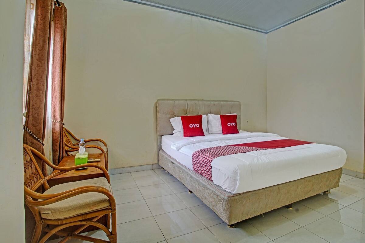 Quarto de hotel com uma cama, uma cadeira de madeira, uma mesa com uma garrafa e uma caixa de lenços, além de uma cortina.