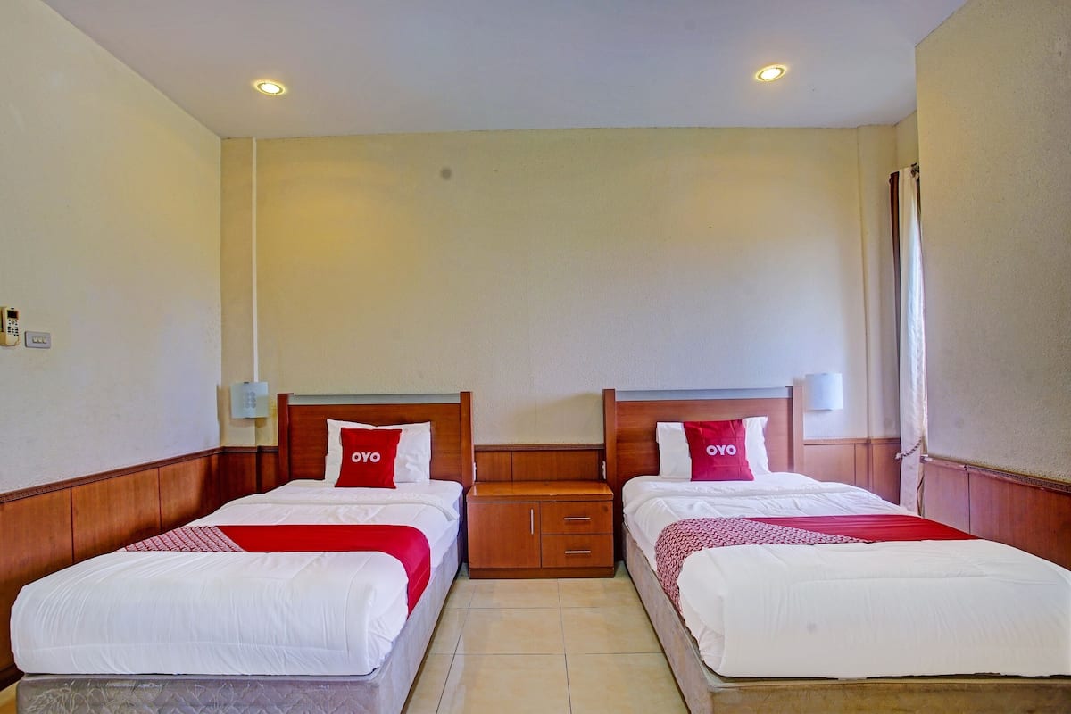 Quarto de hotel com duas camas, cada uma com cabeceira de madeira e roupa de cama vermelha e branca. Há uma mesa de cabeceira de madeira entre as camas, com uma gaveta e uma pequena mesa.