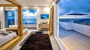 Quarto moderno com cama grande, cabeceira de madeira e vista para o oceano através de amplas janelas.