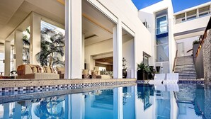 Uma área de piscina moderna com água azul cristalina, um espaço de estar com sofás e uma escadaria que leva a um andar superior.