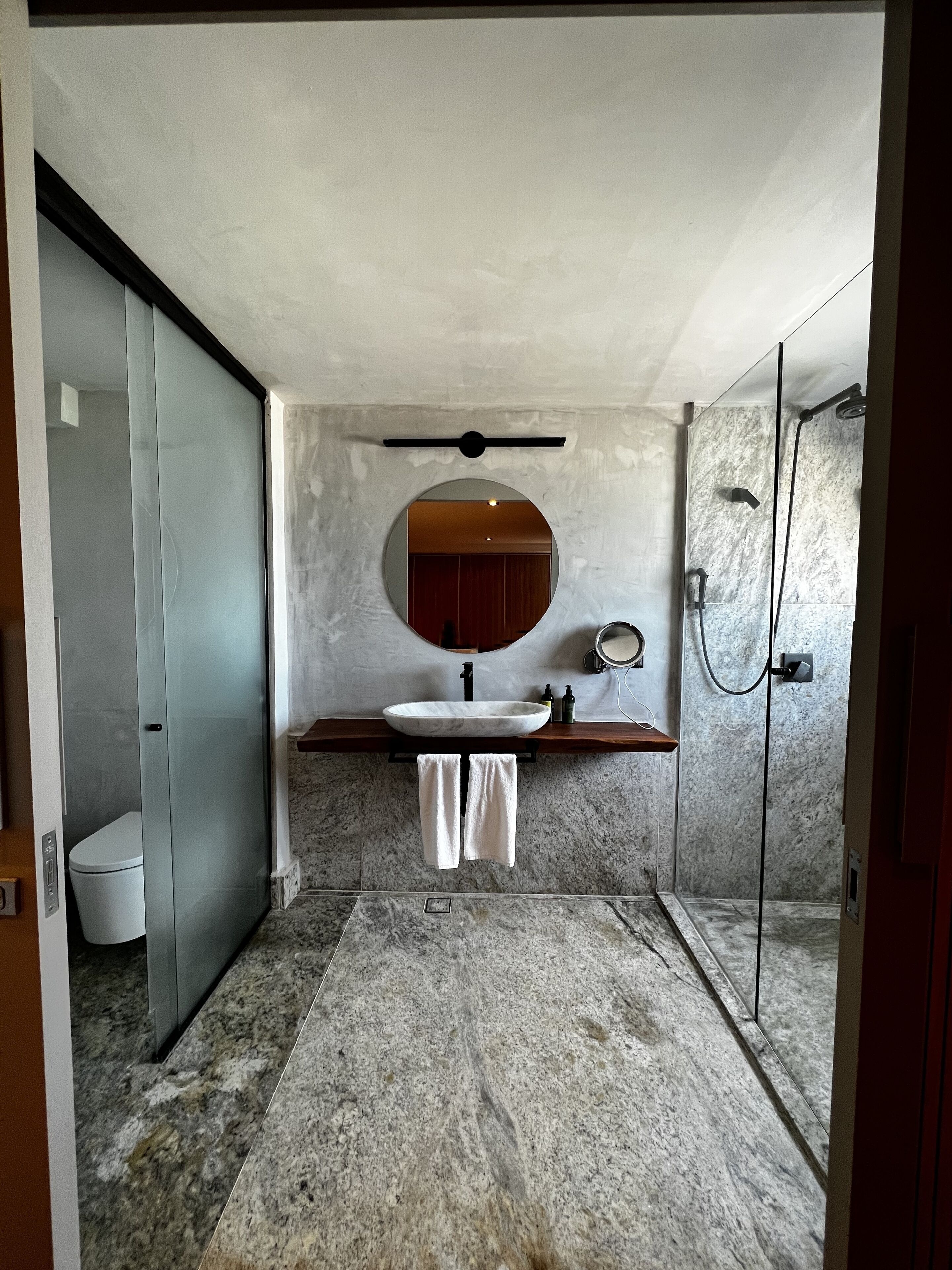 Uma casa de banho moderna com sanita independente, espelho redondo, lavatório com bancada de mármore e cabine de duche com vidro.