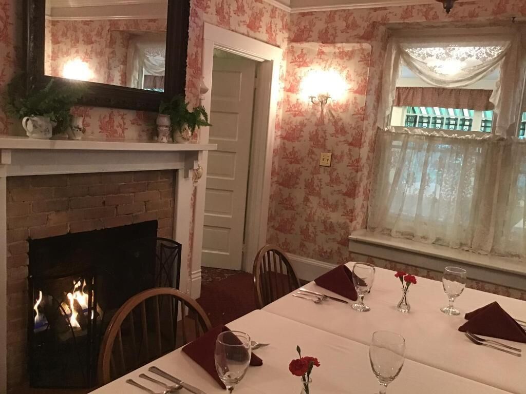 Uma sala de jantar com lareira de tijolos, papel de parede floral e uma mesa posta para quatro pessoas, com taças de vinho e um vaso de rosas vermelhas.