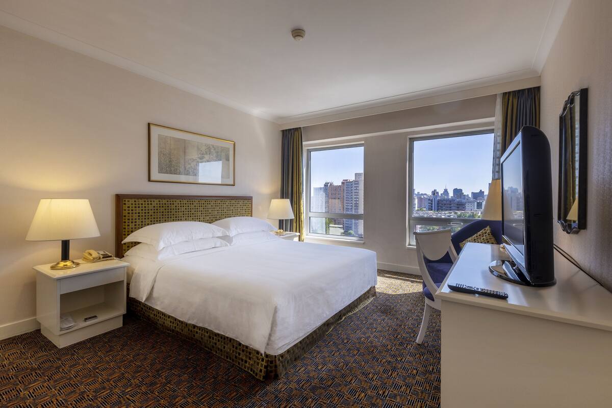 Quarto de hotel com uma cama grande, duas mesas de cabeceira com luminárias, uma escrivaninha com computador e televisão, e vista para a cidade através de amplas janelas.