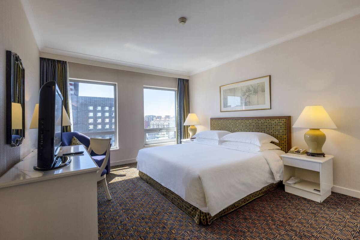 Quarto de hotel com uma cama grande, uma escrivaninha com computador, uma cadeira, mesa de cabeceira com abajur e janela com vista para a cidade.