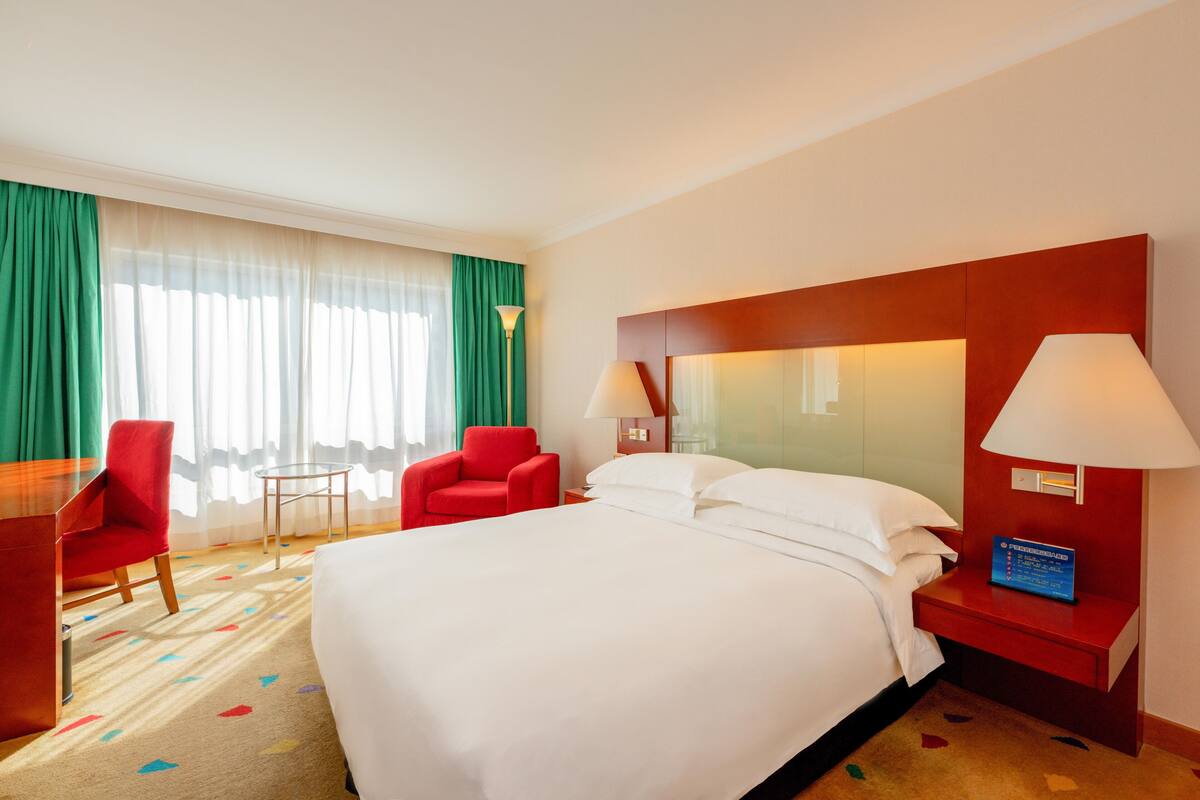 Quarto de hotel com uma cama grande, uma escrivaninha, uma cadeira, uma mesinha e um abajur.