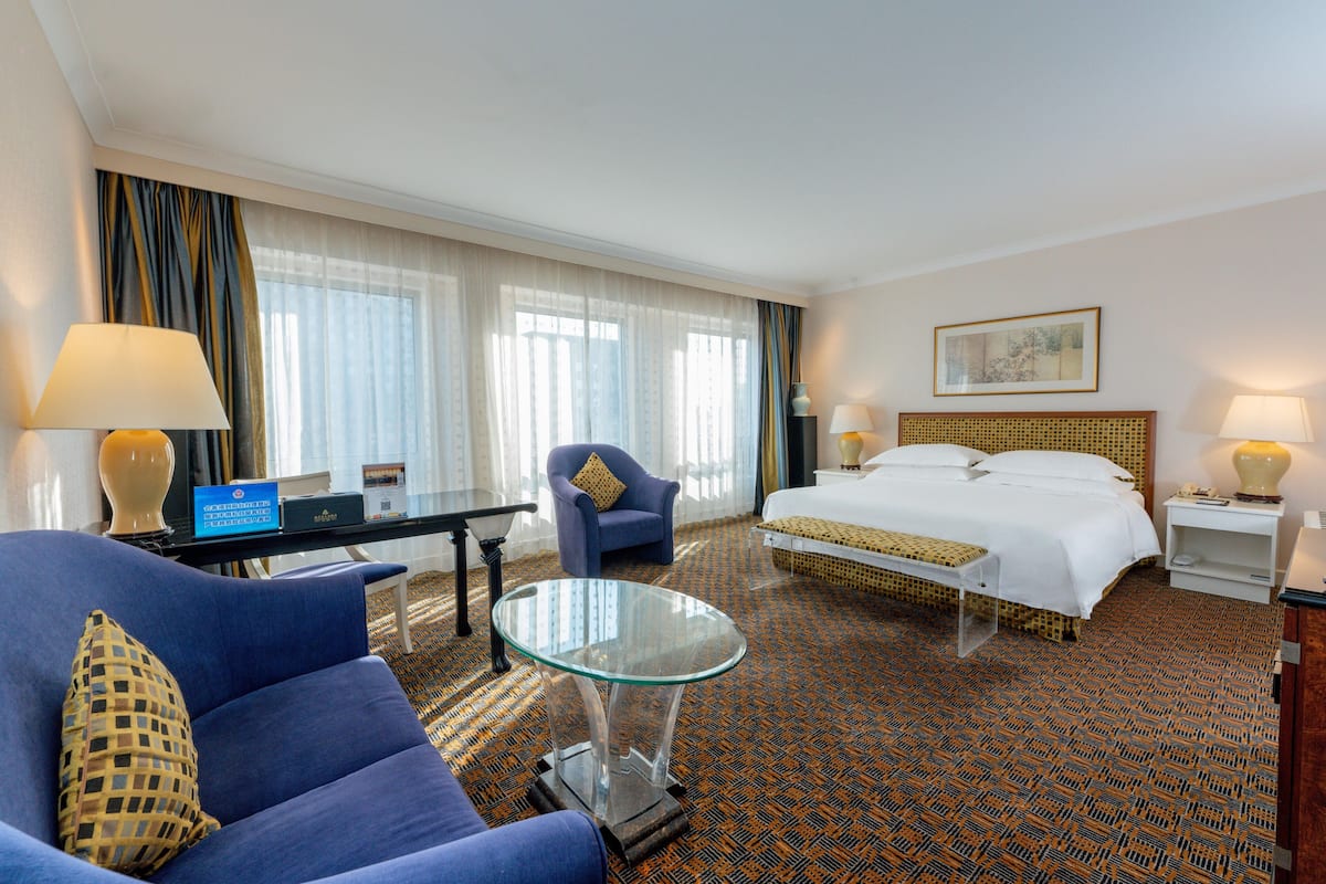 Quarto de hotel com cama, escrivaninha, cadeira, sofá e mesa de centro de vidro.