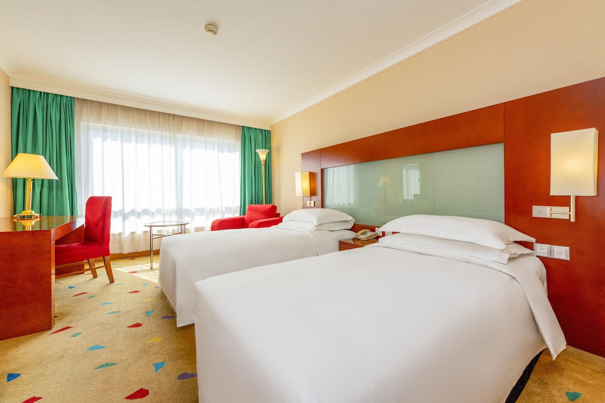 Quarto de hotel com duas camas, uma escrivaninha, uma cadeira, um abajur e uma janela com cortinas.