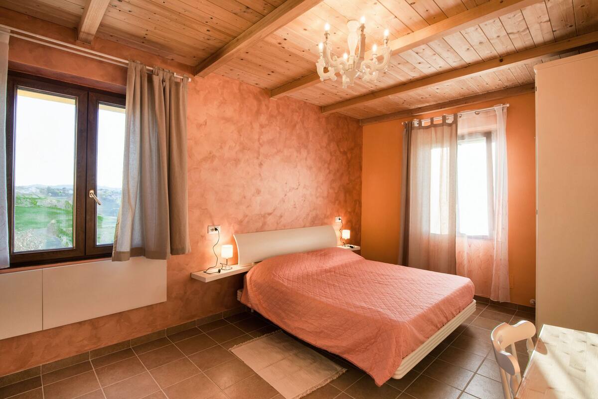 Quarto com cama, mesas de cabeceira, um candelabro e vista para o exterior.