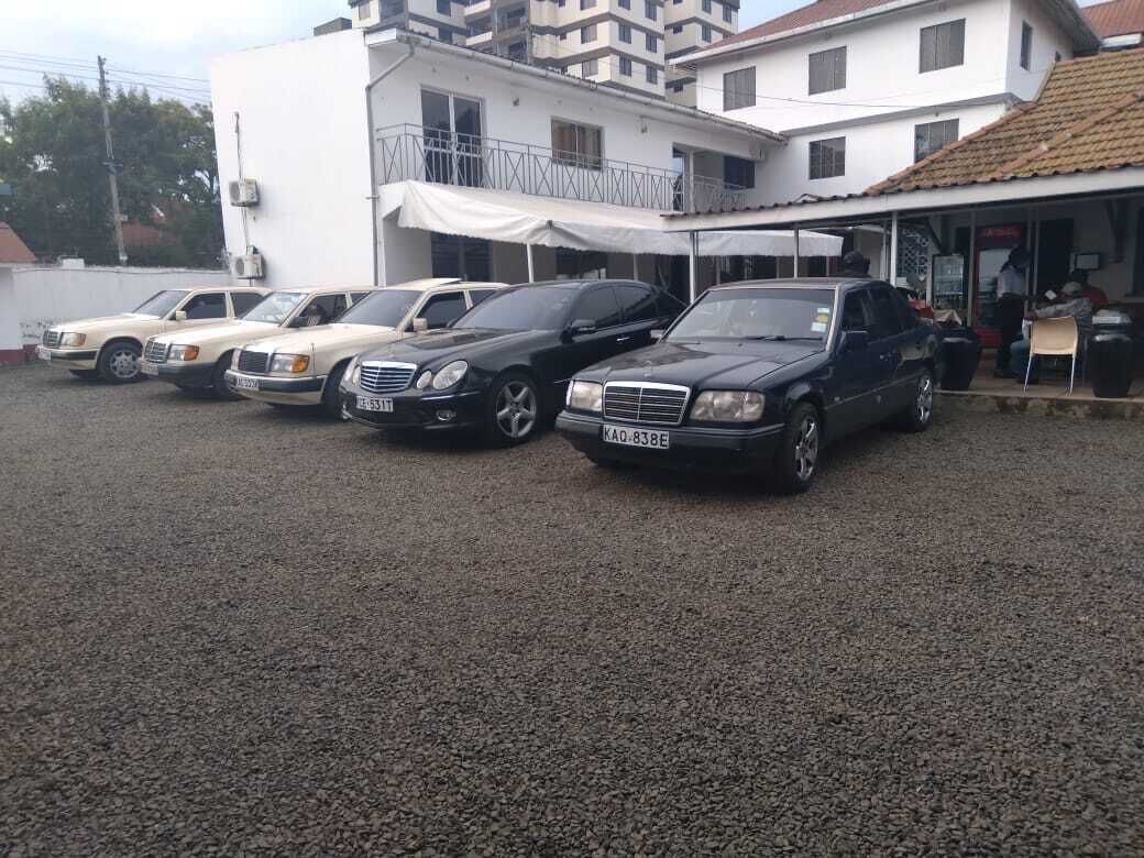 Vários carros estacionados em um estacionamento de cascalho, com edifícios ao fundo.