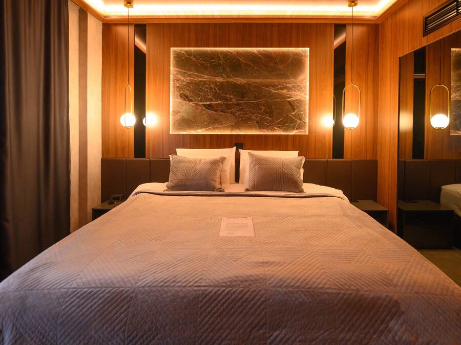Quarto de hotel com uma cama grande, duas mesas de cabeceira, uma luminária fixada na parede e detalhes em painel de madeira.
