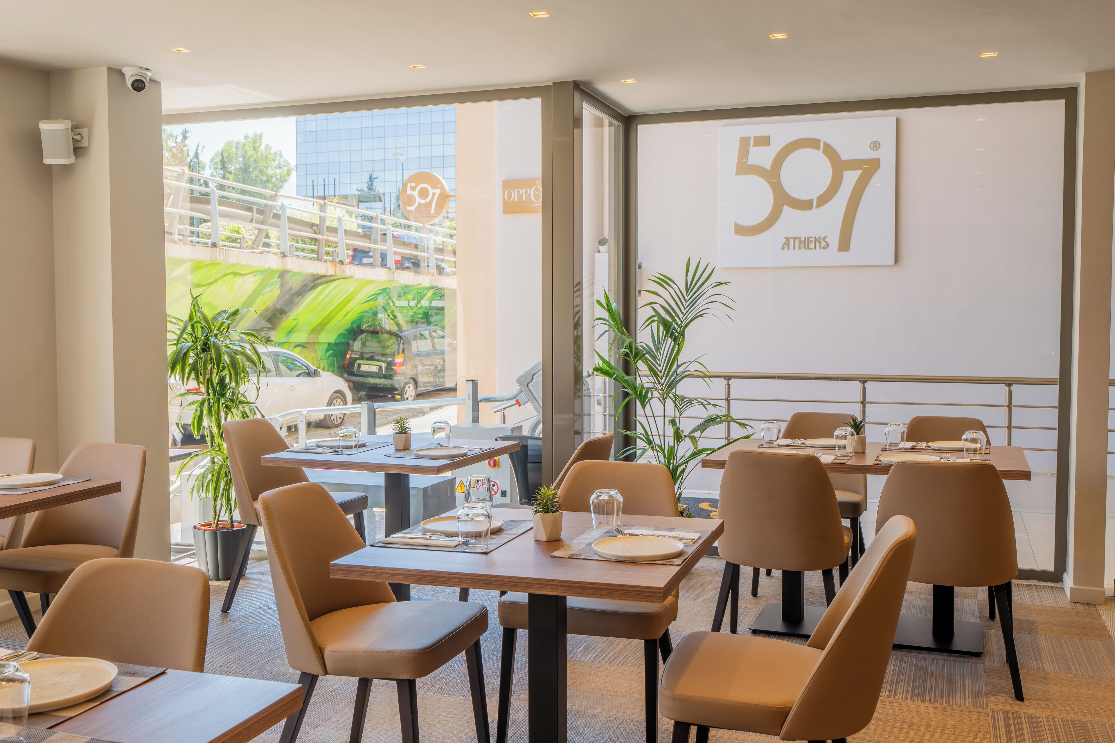 Interior de restaurante moderno com mesas e cadeiras, uma grande janela com vista para a rua, e um letreiro na parede com a inscrição "507 ATHENS".