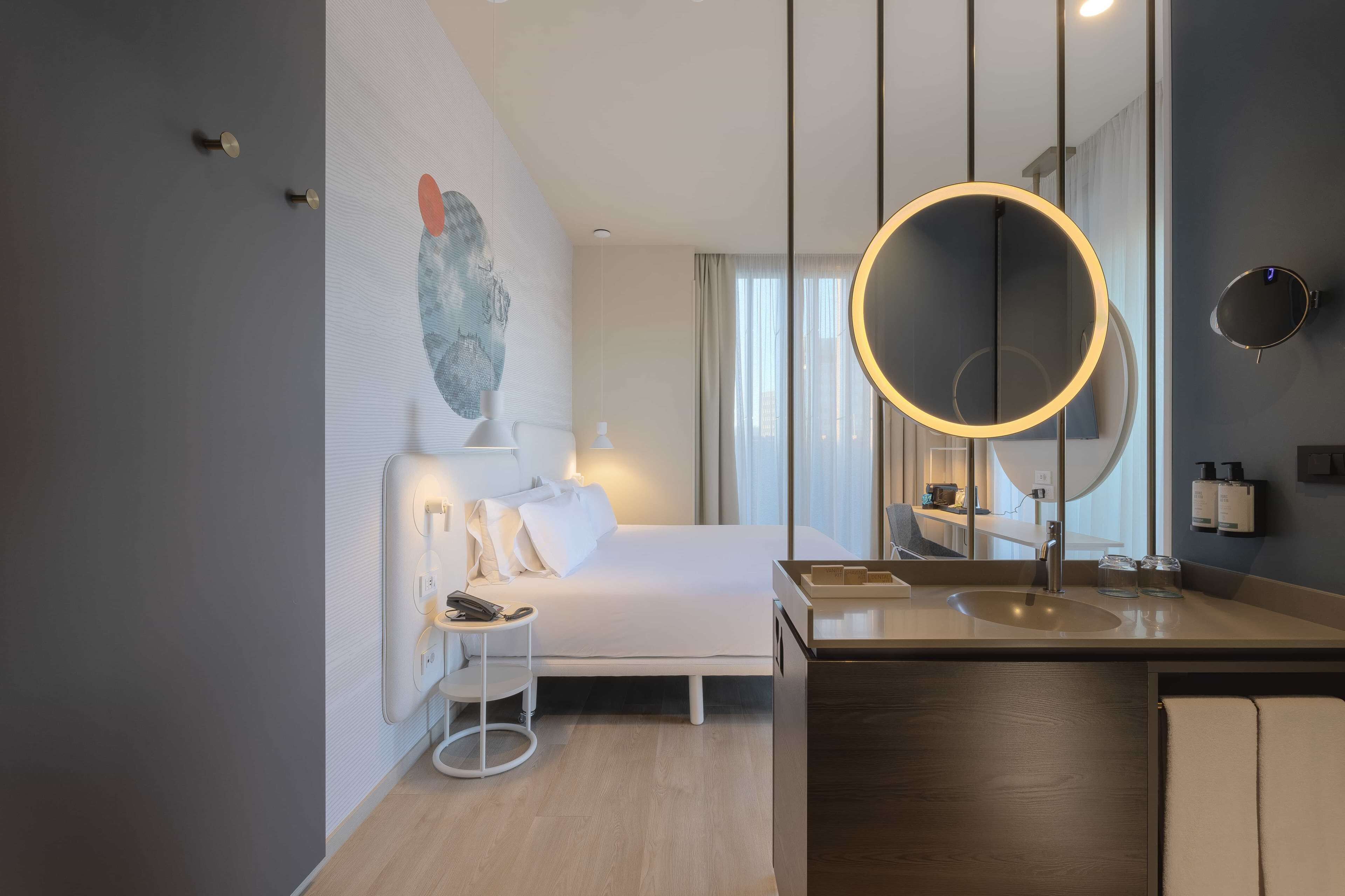 Quarto de hotel moderno com cama, mesa de cabeceira, abajur e um espelho grande com moldura circular.