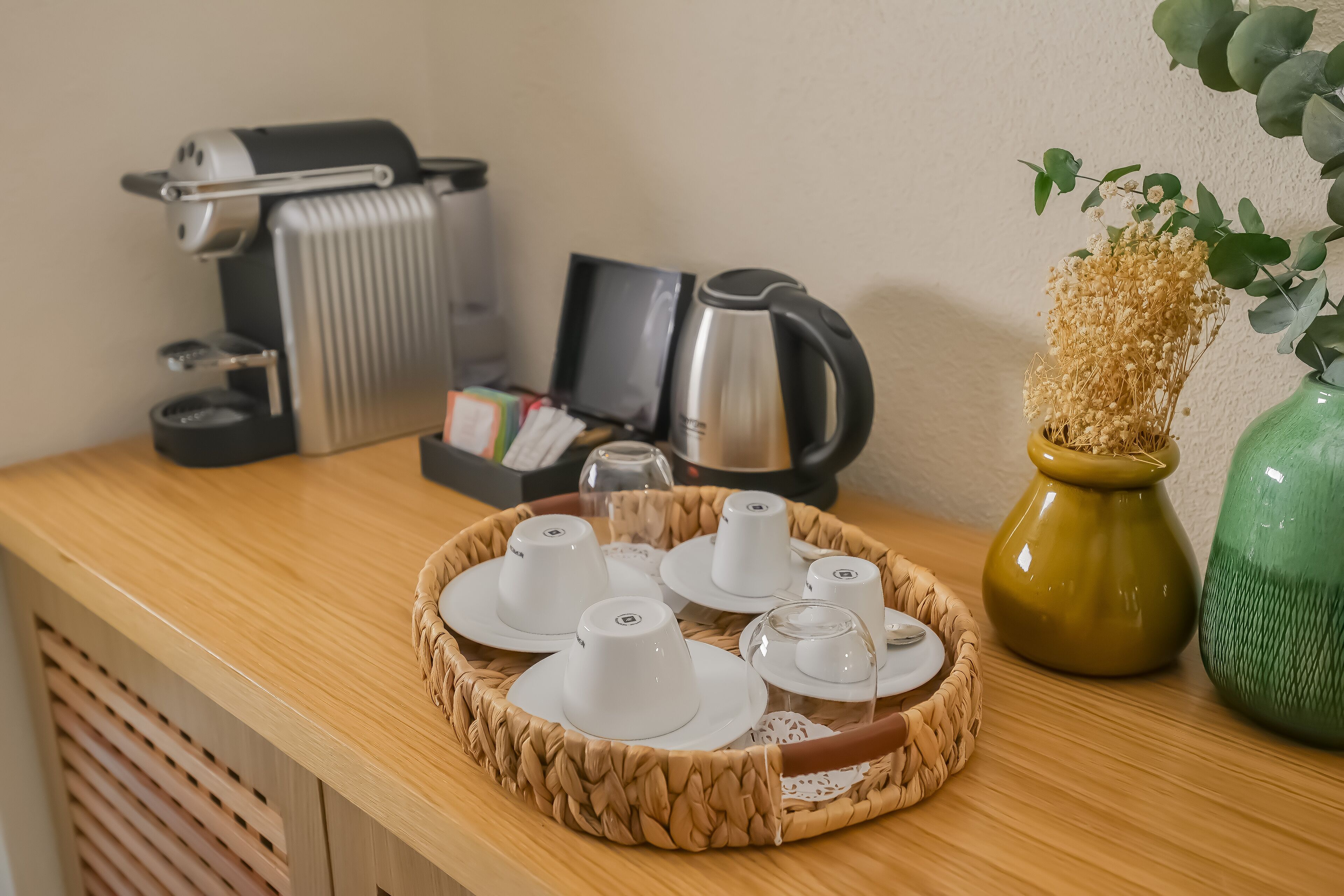 Uma bandeja com xícaras e pires, uma cafeteira, uma chaleira e um vaso com flores secas sobre uma mesa de madeira.