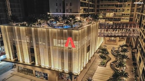 Um hotel moderno com um logo vermelho distinto em formato de 'A', uma área externa coberta com palmeiras e um átrio bem iluminado.