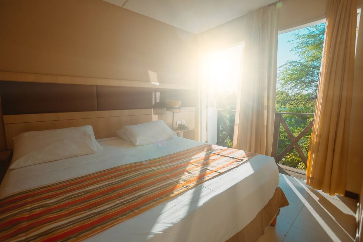 Quarto de hotel com uma cama grande, cabeceira, mesa de cabeceira e janela com cortinas.