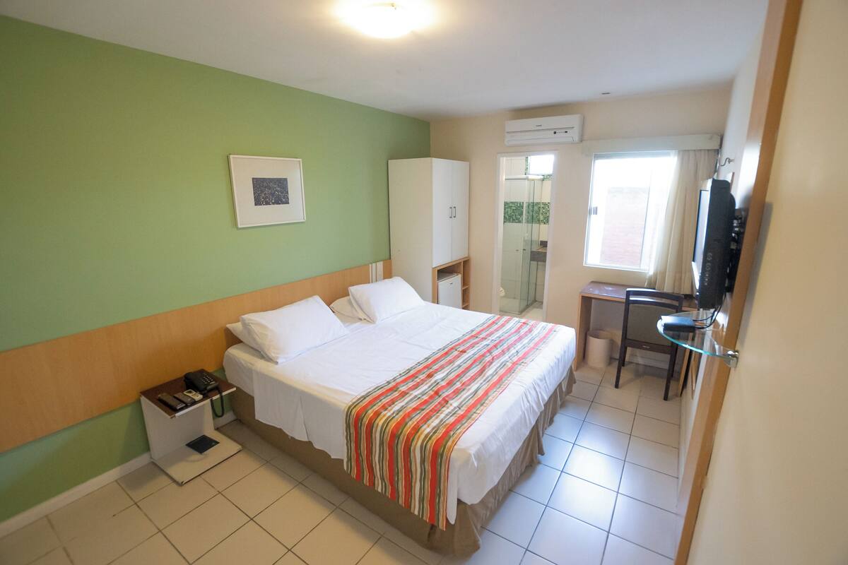 Quarto de hotel com cama, escrivaninha, guarda-roupa e televisão.