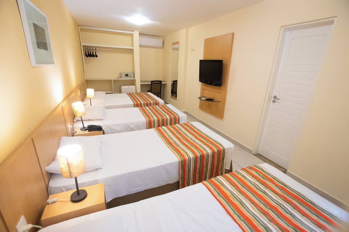 Quarto de hotel com três camas de solteiro, televisão fixada na parede, escrivaninha com cadeira e um cabideiro.