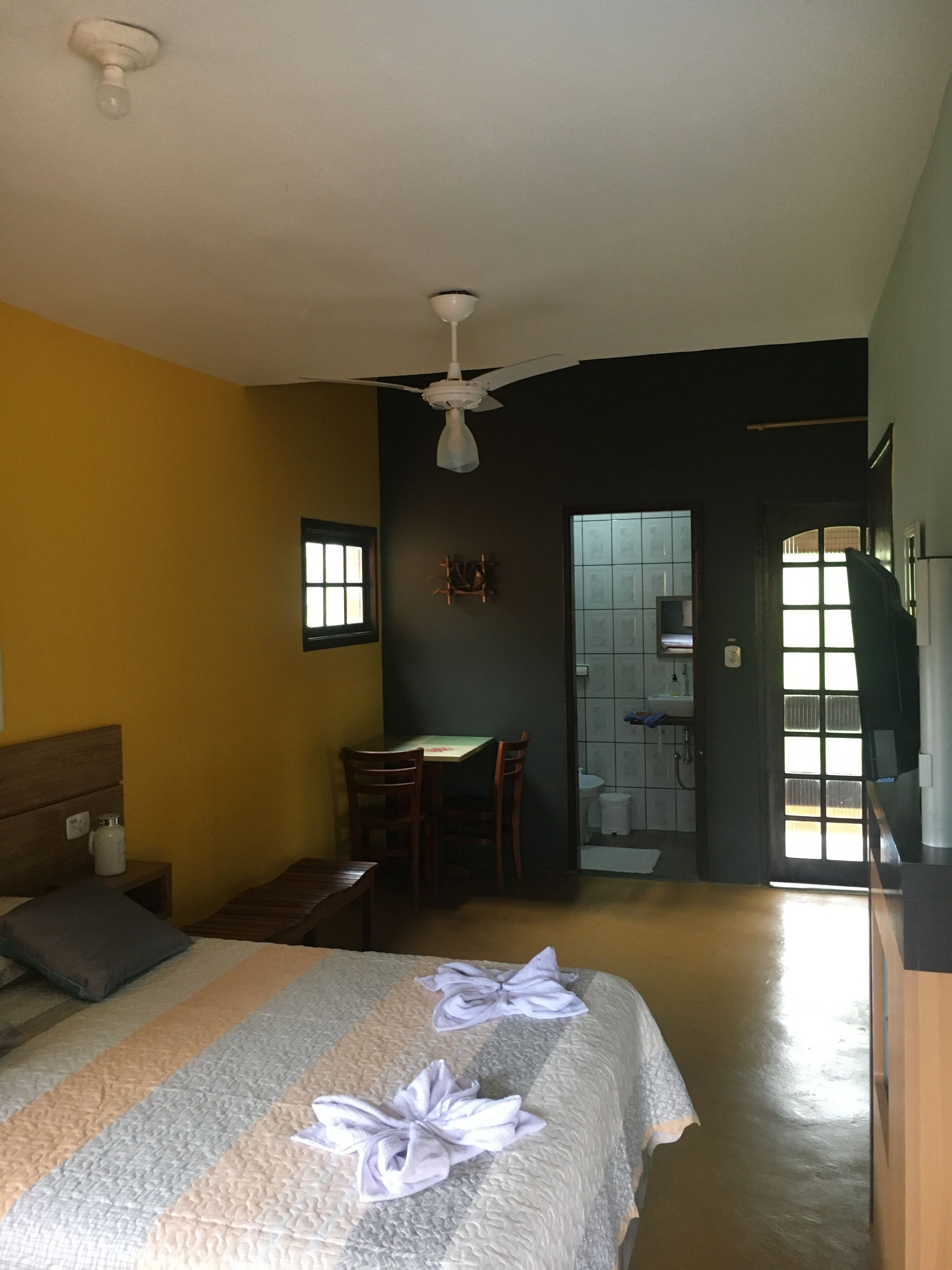Quarto com cama, escrivaninha, cadeira e banheiro visível por uma porta aberta.