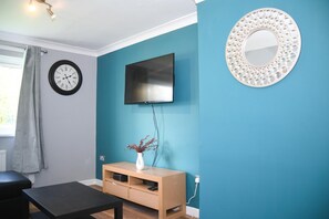 Quarto com parede verde-água, televisão montada, espelho redondo, relógio e armário de madeira com um vaso.