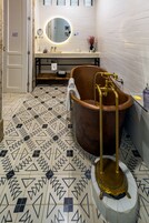 Um banheiro com uma grande banheira de cobre, um espelho redondo e um chão de azulejos brancos com padrões geométricos.