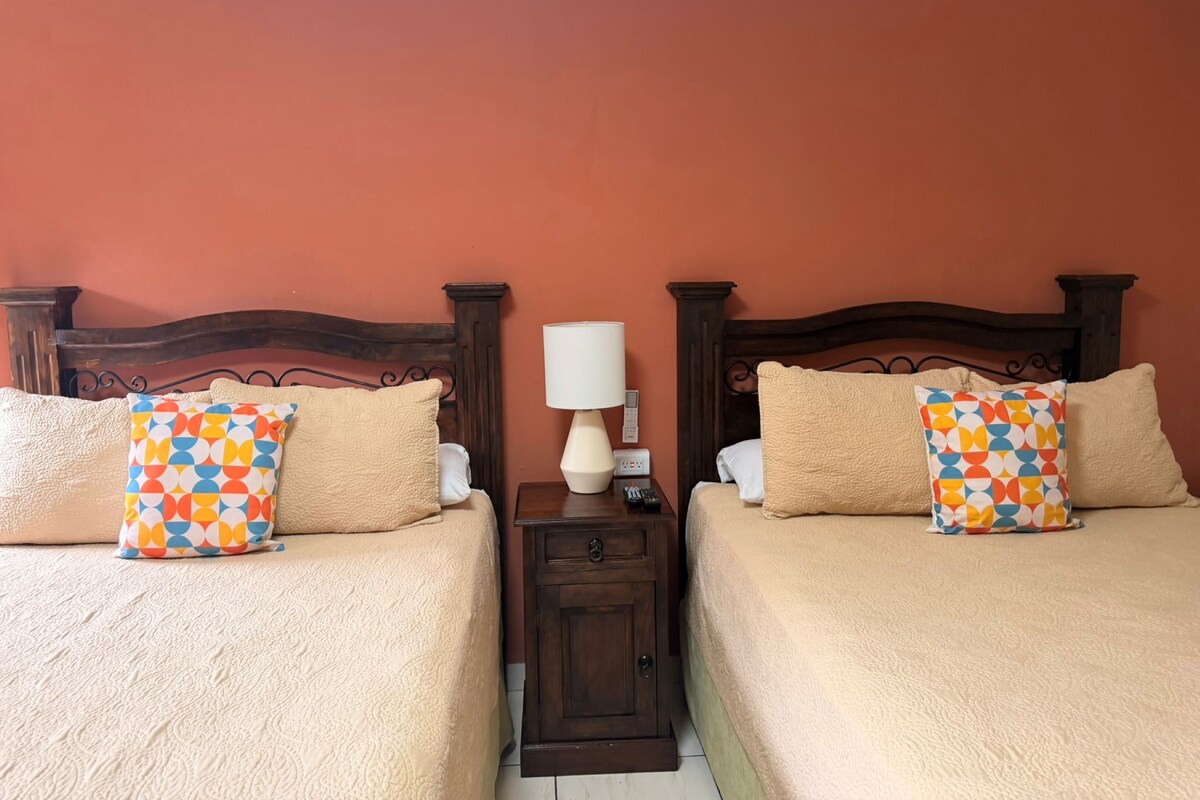 Quarto de hotel com duas camas, cada uma com almofada estampada, mesa de cabeceira com abajur e criado-mudo com telefone.