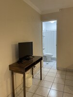 Quarto com suporte de TV de madeira, um balde branco e banheiro visível através de uma porta aberta.