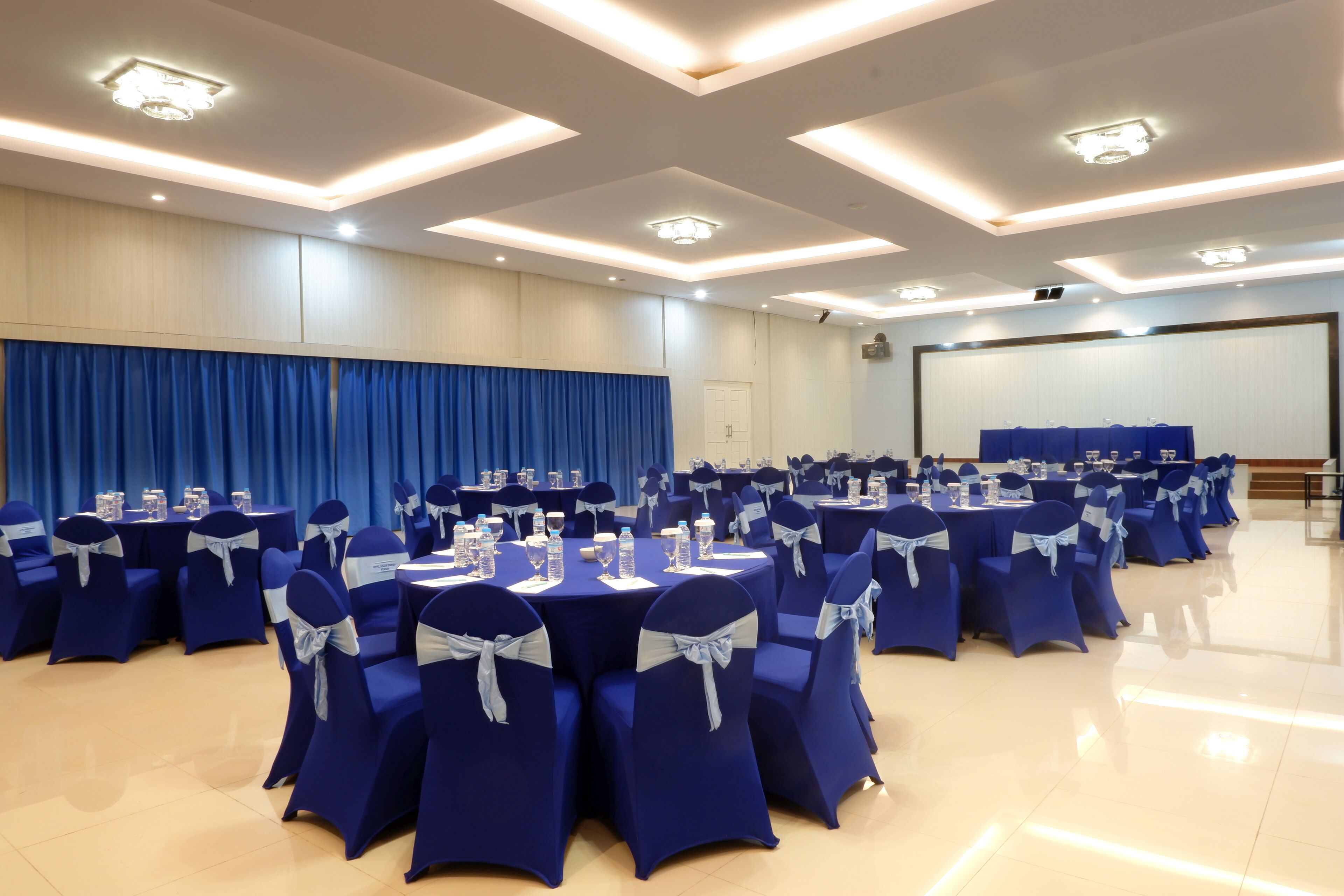 Sala de conferências com fileiras de mesas e cadeiras, cada conjunto com garrafas de água e copos, e uma tela grande na frente.
