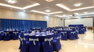 Sala de conferências com fileiras de mesas e cadeiras, cada conjunto com garrafas de água e copos, e uma tela grande na frente.